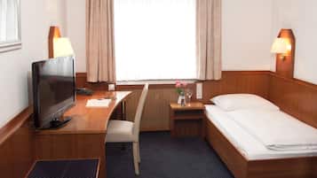 Quarto de hotel com cama, secretária com televisão, cadeira, mesinha com abajur e janela com cortinas.