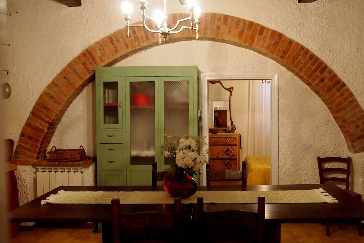 Uma sala de jantar com arco de tijolos, um armário verde, um vaso com flores e um lustre.