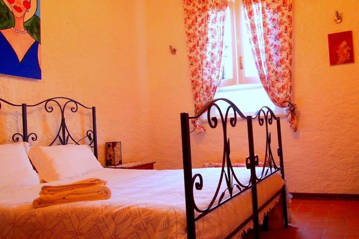 Quarto com cama, mesa de cabeceira, janela com cortinas, um quadro na parede e uma porta.