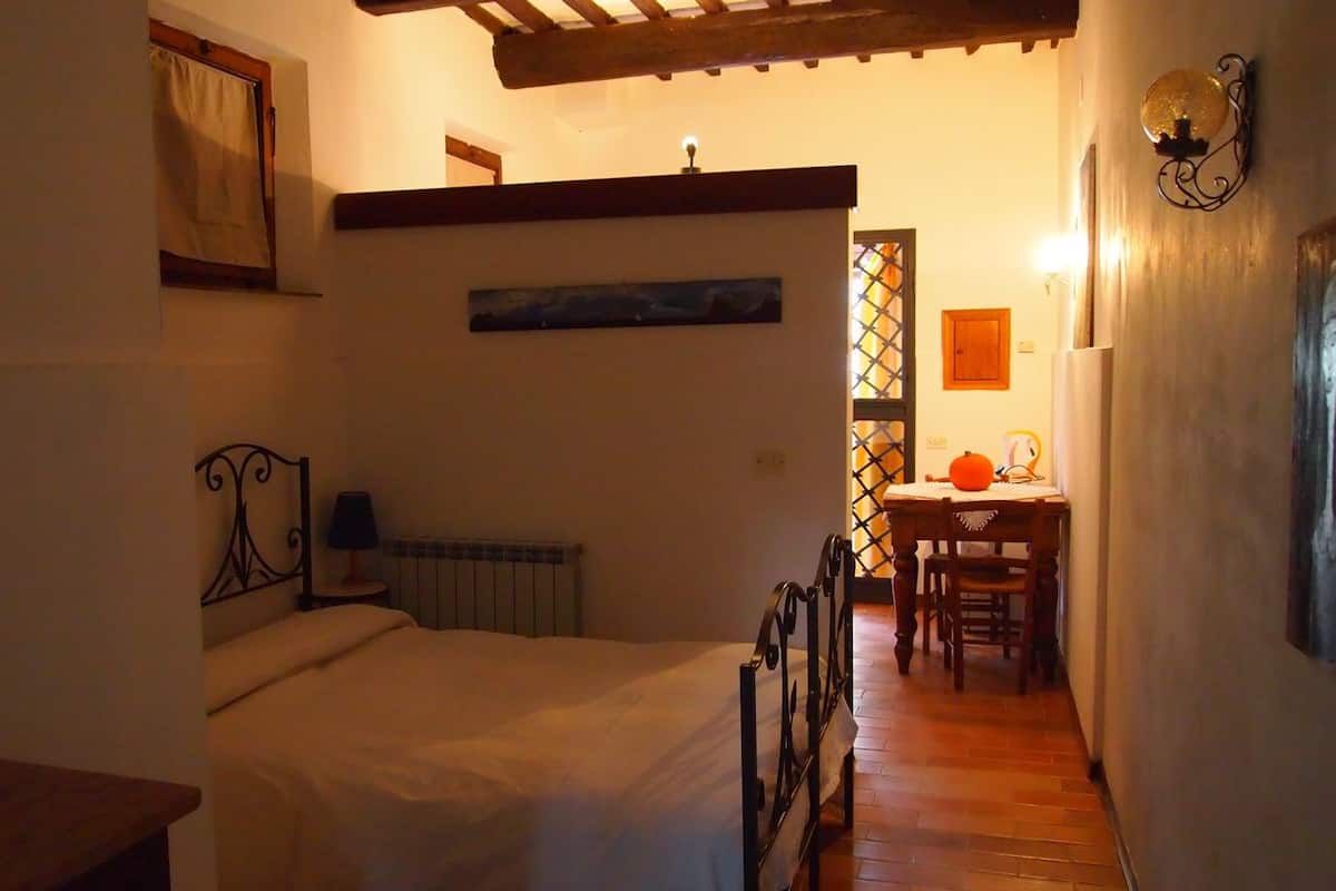 Quarto com cama, mesa de cabeceira, janela com cortinas, teto de madeira e uma pequena mesa com cadeira.