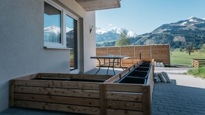 Um deck de madeira com mesa e cadeiras, uma janela e vista para montanhas cobertas de neve.