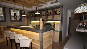 Uma área de bar com banquetas de madeira, uma ilha central com pia e diversas garrafas e copos nas prateleiras.