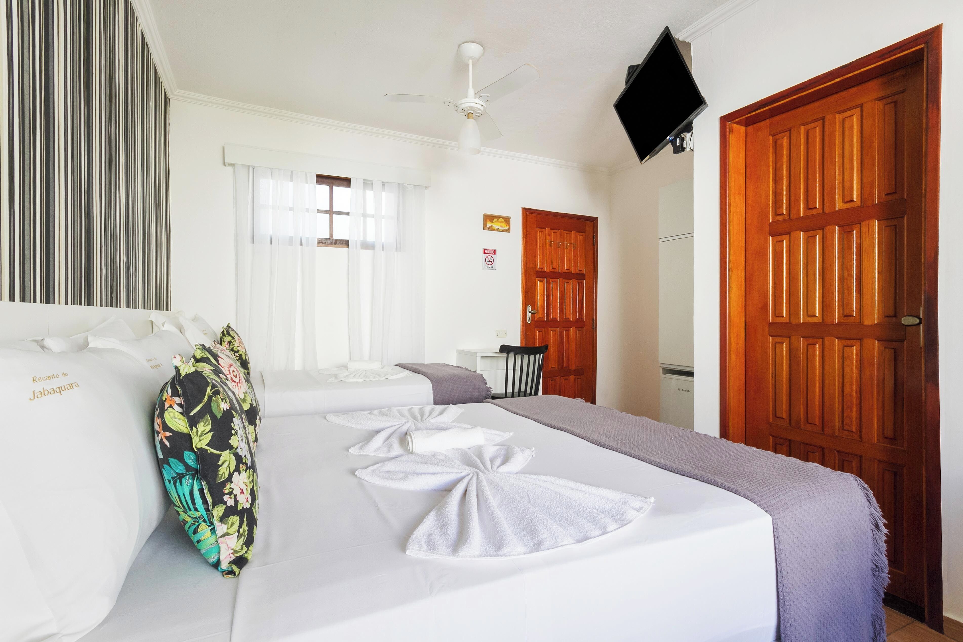 Quarto de hotel com duas camas, um ventilador de teto, uma televisão fixada na parede e uma porta de madeira.