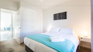 Quarto de hotel bem arrumado com uma cama, uma mesa de cabeceira com abajur, uma obra de arte na parede e uma porta que dá para outro quarto.