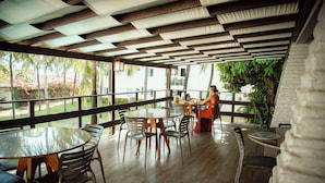 Um espaço de refeições interno com mesas e cadeiras de madeira, vista para o exterior e uma pessoa sentada à mesa.
