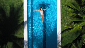 Uma piscina retangular com água azul cristalina, rodeada por um gramado bem cuidado e palmeiras.