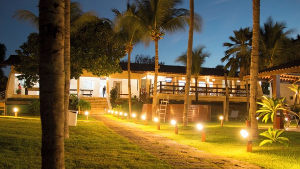 Um hotel com entrada bem iluminada, palmeiras e um caminho pavimentado até o edifício.
