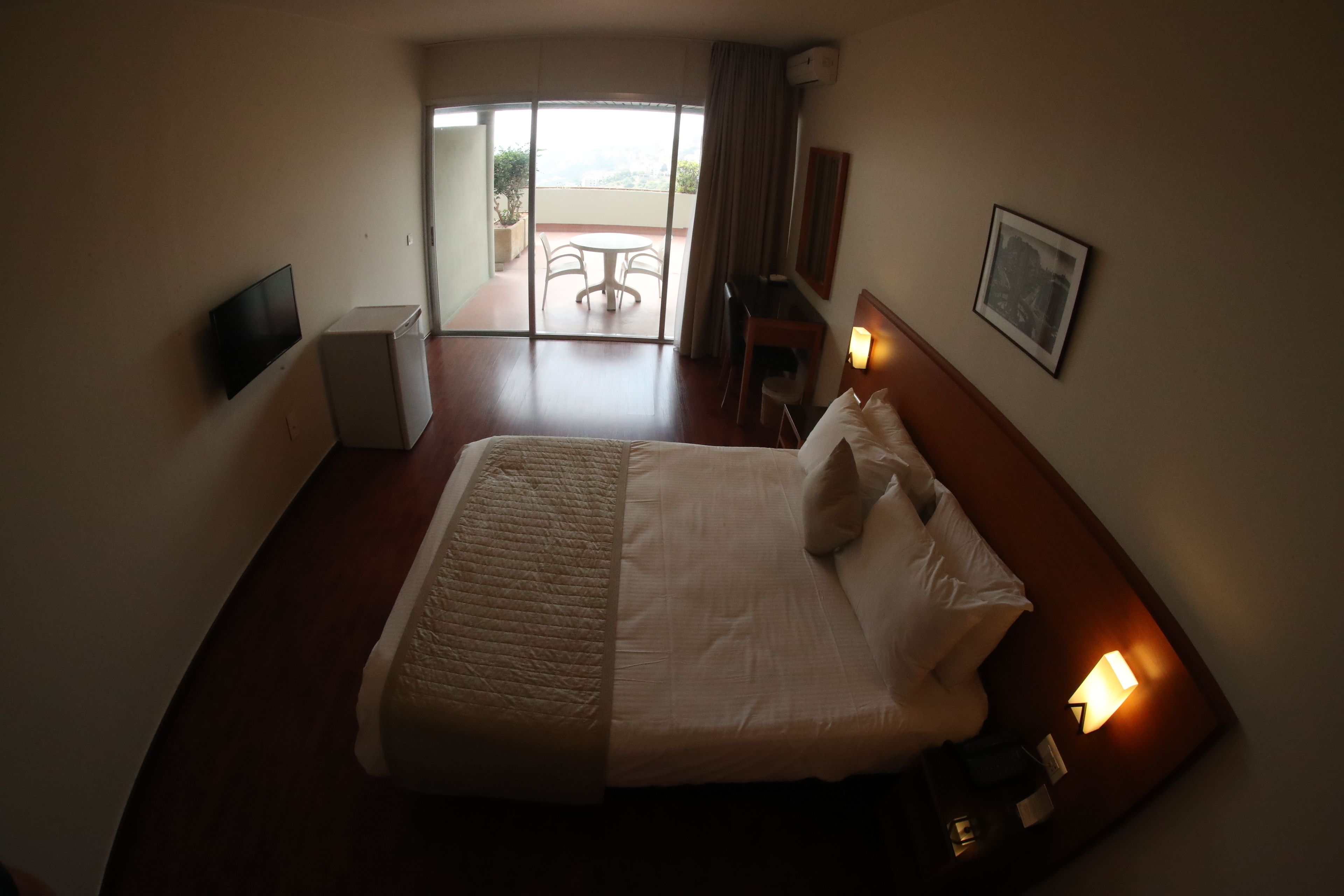 Quarto de hotel com cama, escrivaninha, poltrona, televisão e varanda com mesa e cadeiras.