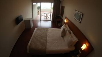 Quarto de hotel com cama, escrivaninha, poltrona, televisão e varanda com mesa e cadeiras.