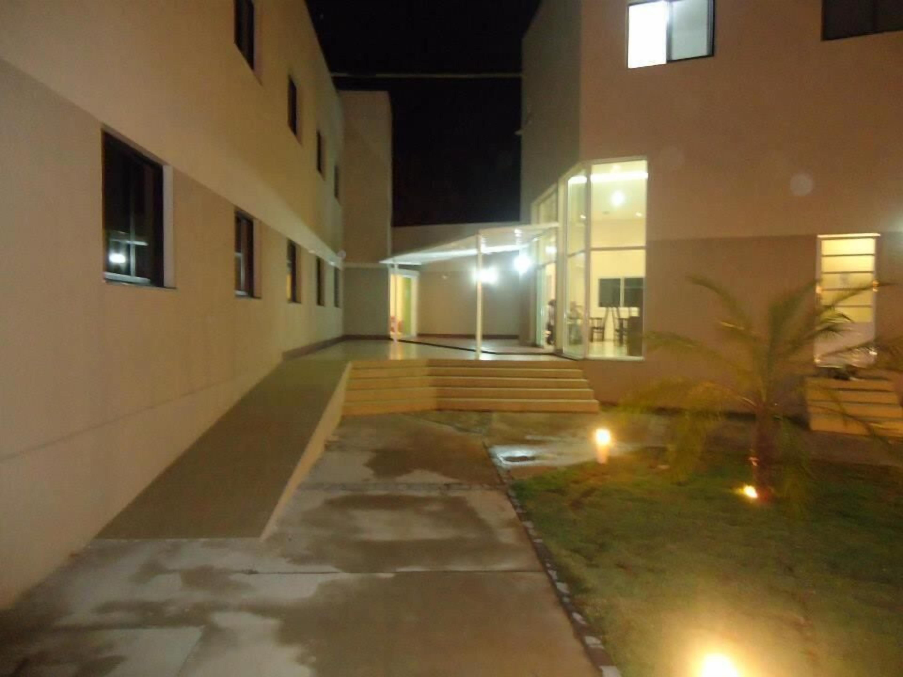 Um edifício com área externa coberta, degraus que levam a uma porta de vidro e um pequeno jardim com uma palmeira.