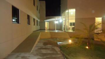 Um edifício com área externa coberta, degraus que levam a uma porta de vidro e um pequeno jardim com uma palmeira.