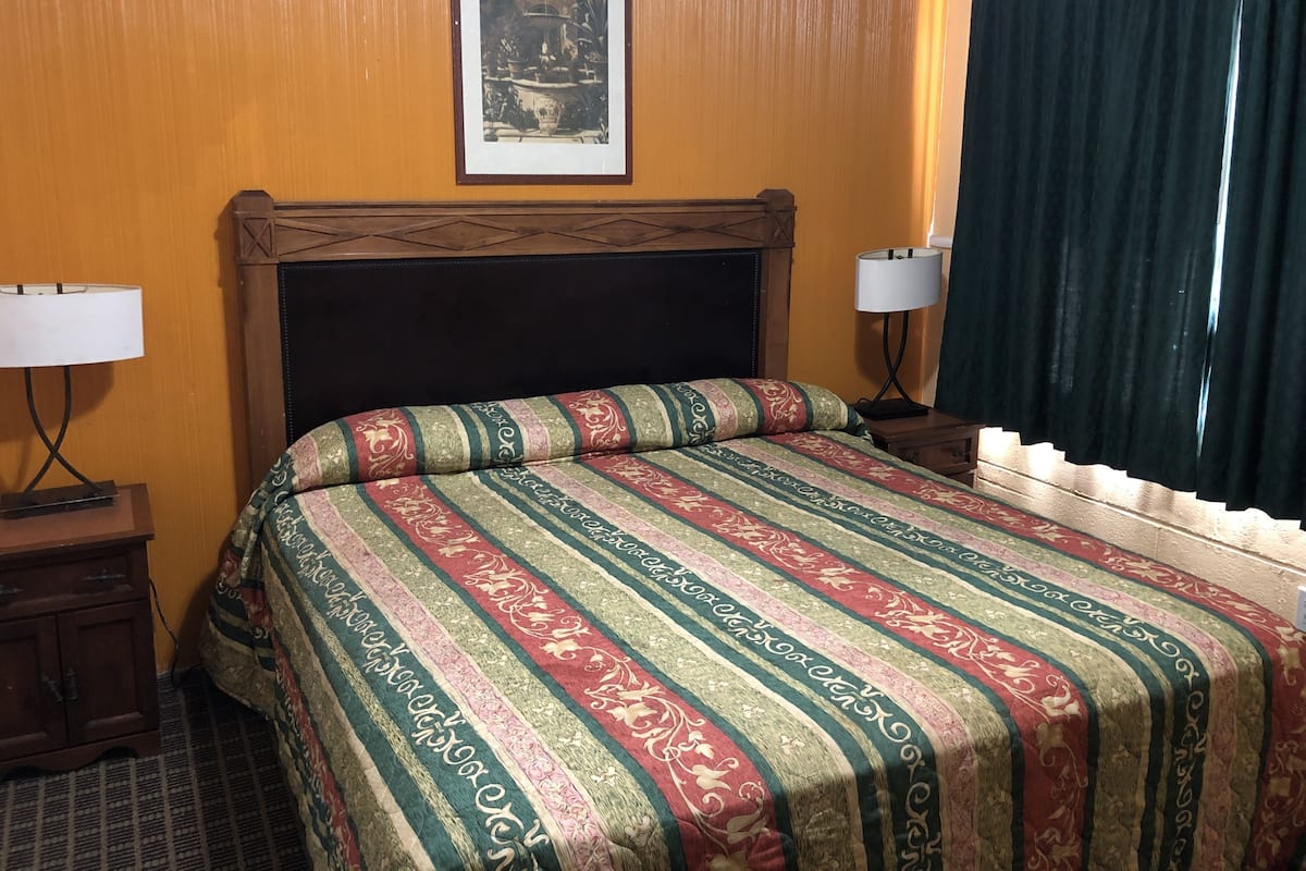 Quarto de hotel com uma cama grande, duas mesas de cabeceira com luminárias, um quadro na parede e uma janela com cortinas.