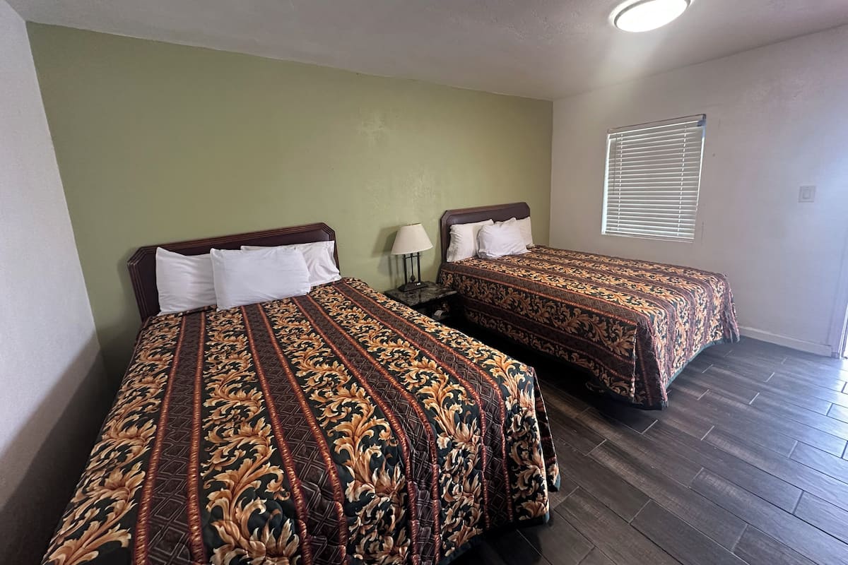 Duas camas com roupa de cama estampada, uma mesa de cabeceira com abajur, uma janela com persianas e uma luminária no teto.