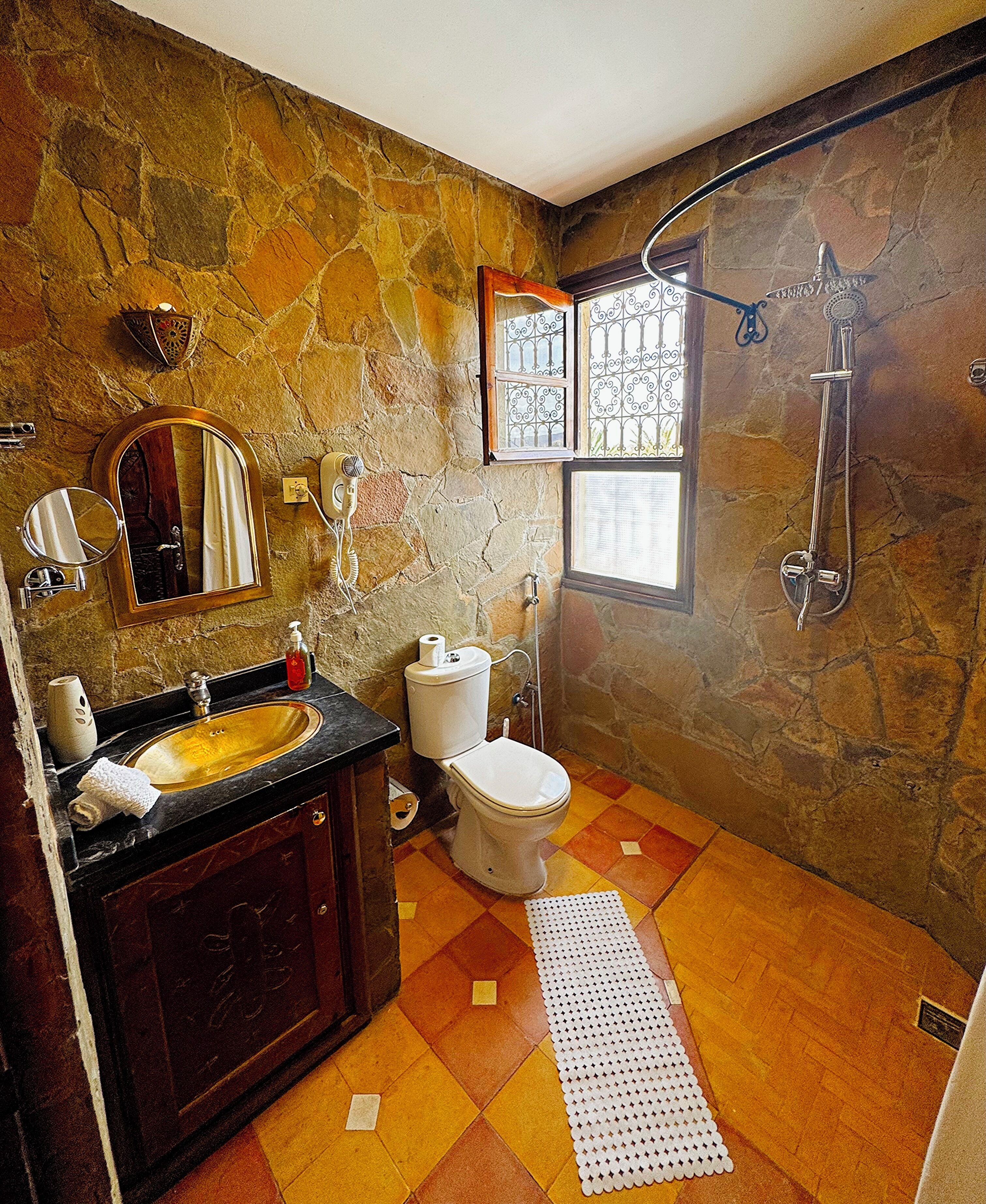 Casa de banho com parede de pedra, pia, sanita, chuveiro e janela com grades de ferro decorativas.