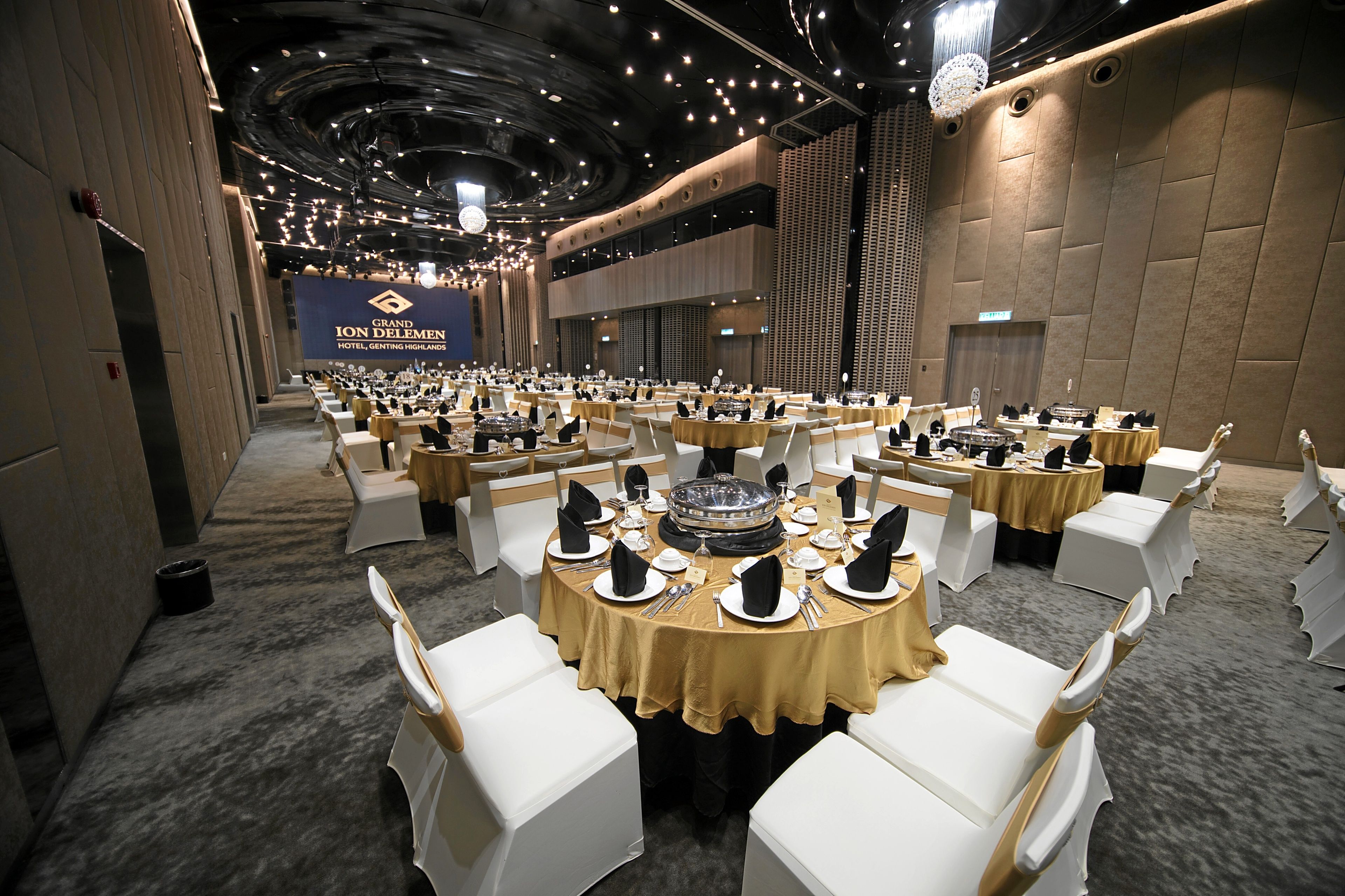 Uma sala de banquetes com mesas redondas preparadas para um evento formal, com cadeiras brancas e toalhas de mesa douradas.