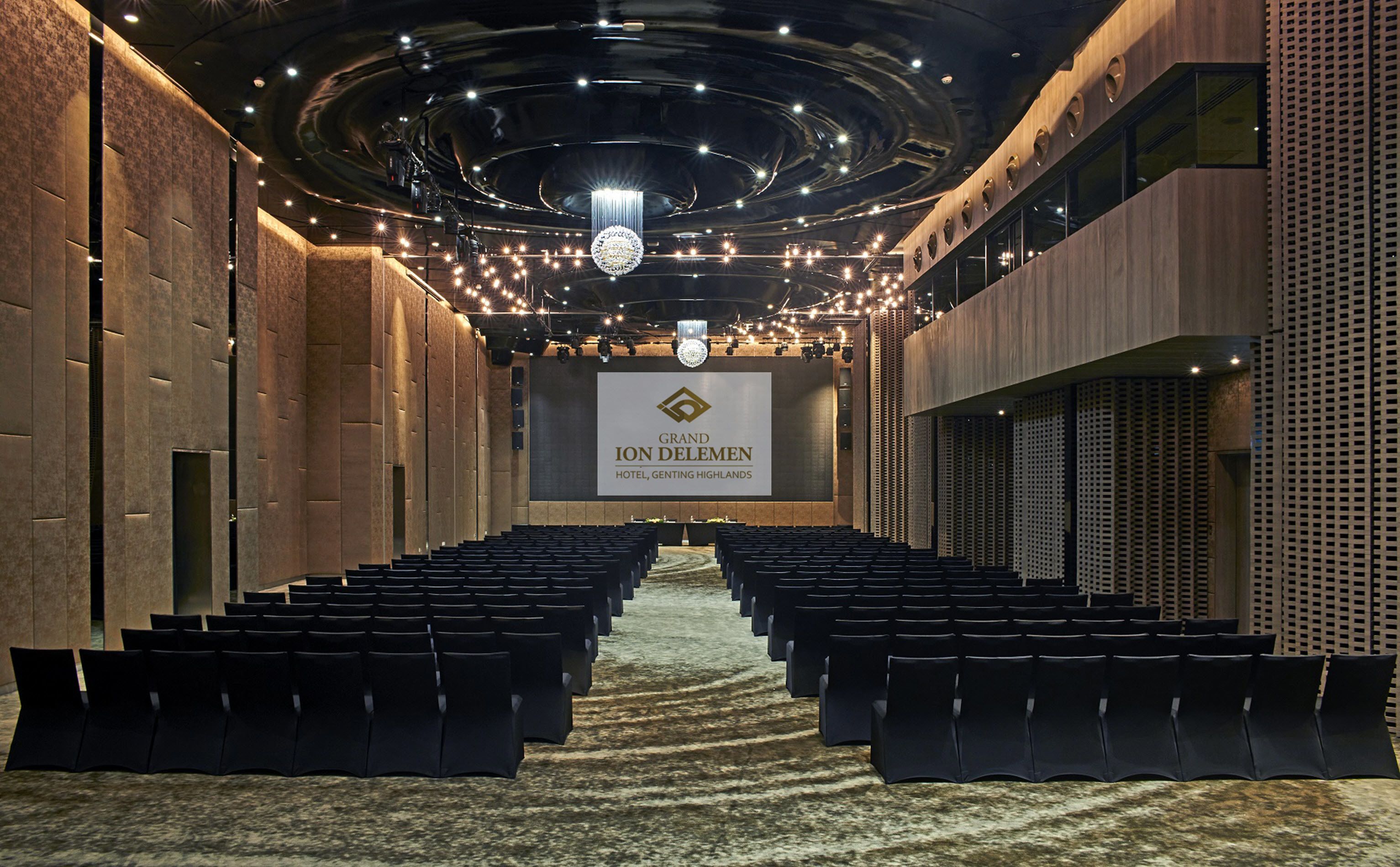 Uma sala de conferências com fileiras de cadeiras pretas, uma tela grande exibindo o logotipo do Grand Ion Delemen Hotel e um design de teto moderno.