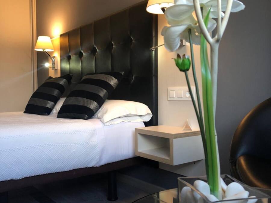 Quarto de hotel moderno com cabeceira estofada, cama com roupa de cama branca e almofadas brancas e pretas, mesas de cabeceira com abajures, e um vaso de vidro transparente com pedras brancas e uma orquídea branca.