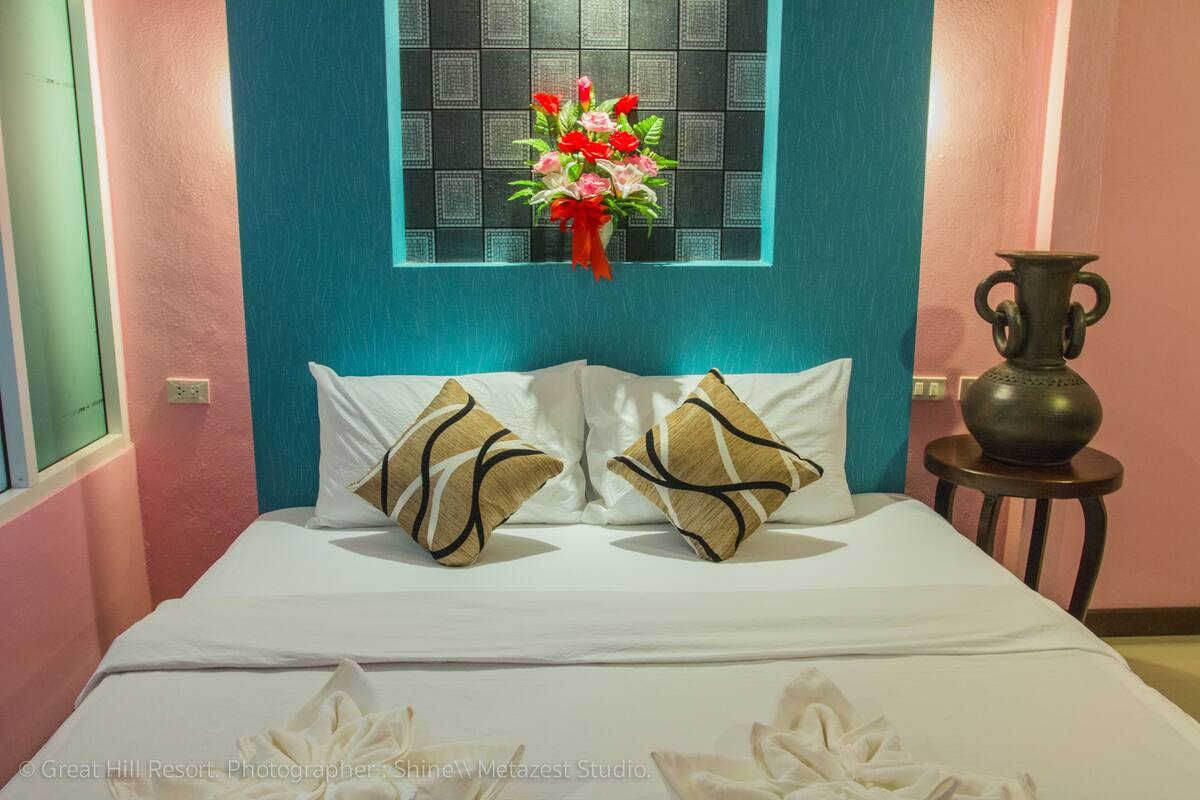 Cama bem arrumada com lençóis brancos e duas almofadas decorativas, um vaso com flores sobre uma mesinha ao lado e uma janela com vista decorativa.