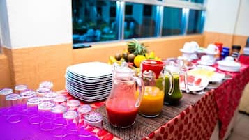 Uma mesa de buffet com diversos alimentos e bebidas, incluindo um jarro de suco vermelho, um jarro de suco verde e uma pilha de pratos.