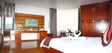 Quarto de hotel com secretária de madeira, televisão, cama com travesseiro em forma de cisne e uma janela grande com cortinas.