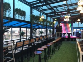 Um bar no terraço com fileiras de banquetas de madeira, um palco com iluminação colorida e plantas penduradas.
