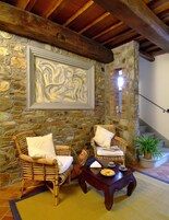 Um quarto com paredes de pedra, duas cadeiras de vime e uma mesa de centro de madeira, um quadro abstrato emoldurado, e uma escada que leva a uma varanda.