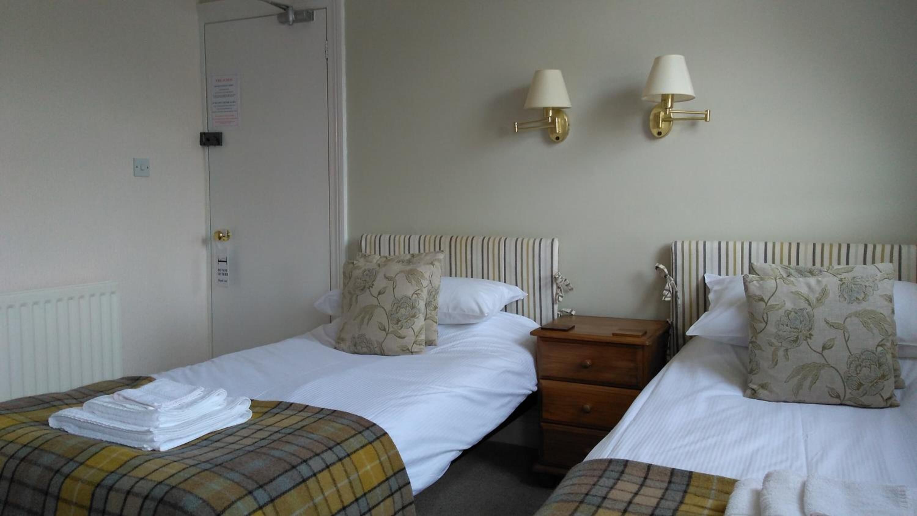 Quarto de hotel com duas camas de solteiro, mesa de cabeceira, radiador e duas luminárias fixadas na parede.