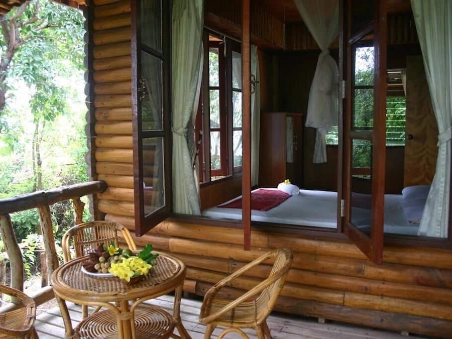 Uma cabana de madeira com varanda, móveis de vime e uma cama com travesseiro vermelho.