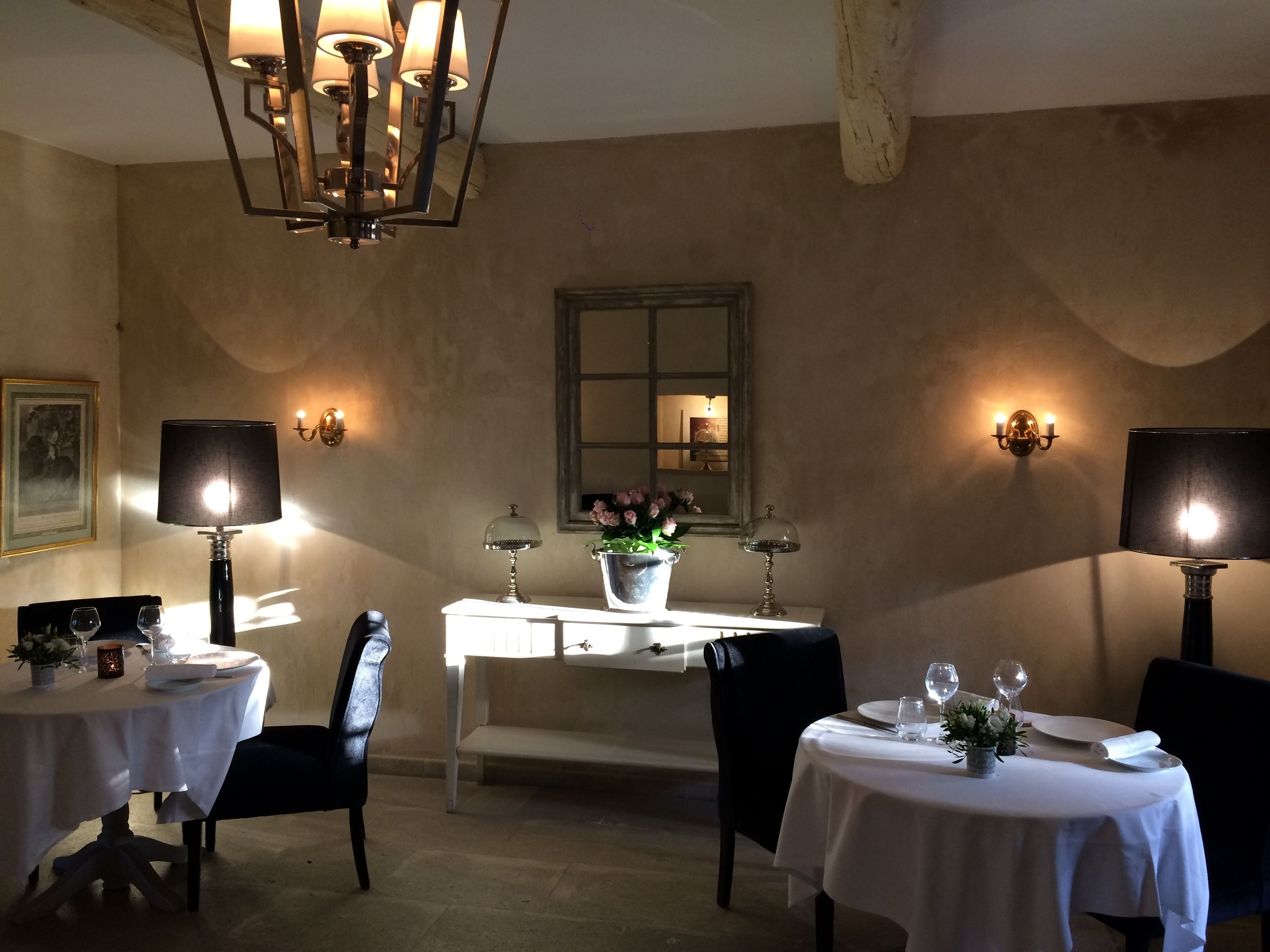 Uma sala de jantar com duas mesas, cada uma com toalha branca, cadeiras pretas e um candelabro. Há arandelas e um espelho emoldurado nas paredes.