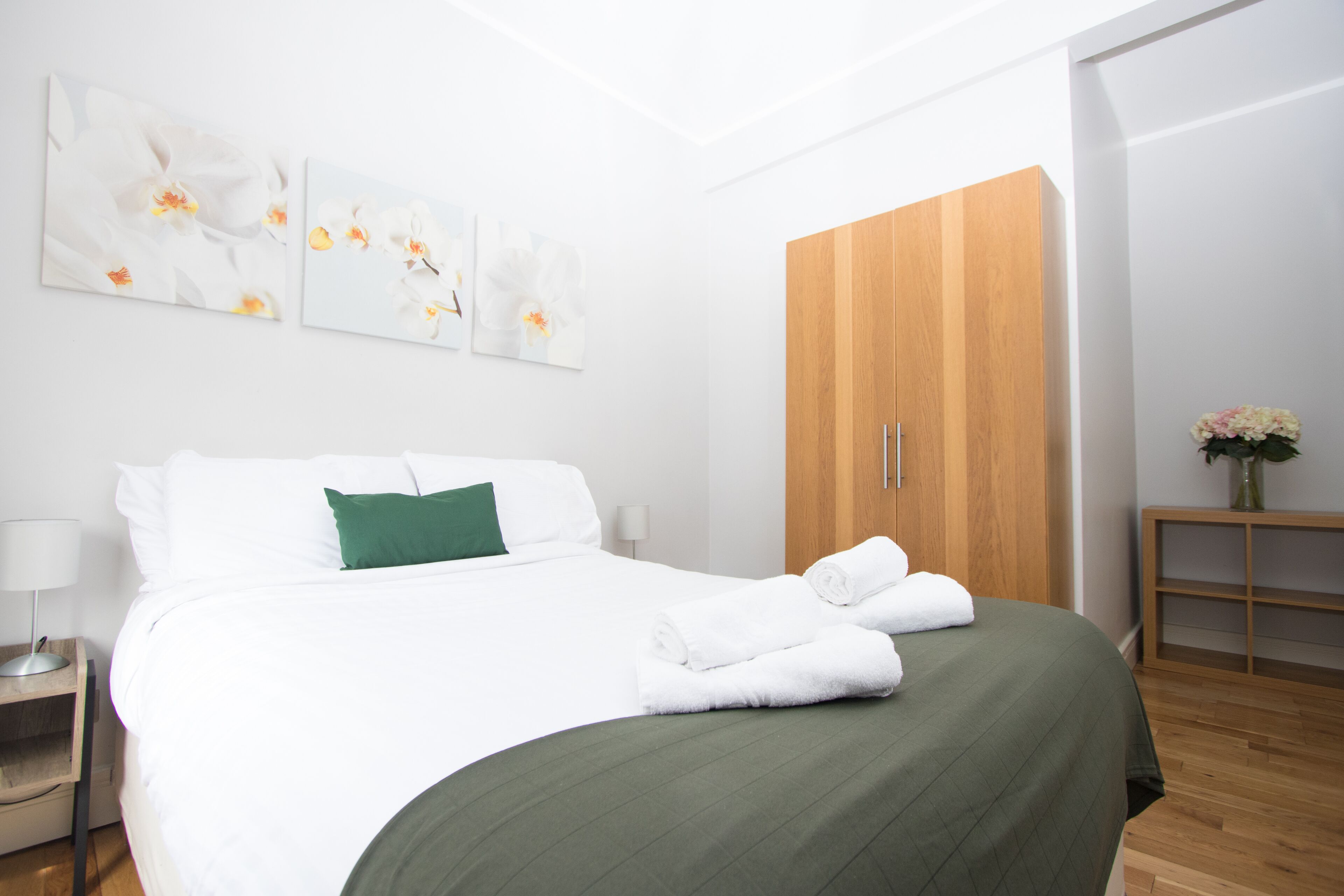 Uma cama bem arrumada com lençóis brancos e um travesseiro verde, um guarda-roupa de madeira e uma pequena mesa de cabeceira com uma luminária.