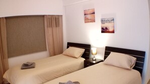 Quarto de hotel com duas camas de solteiro, mesa de cabeceira com abajur e janela com cortinas.