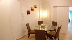 Uma sala de jantar com mesa de vidro, cadeiras ao redor, uma tigela de frutas e um abajur.