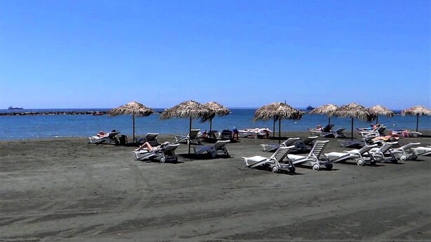Uma praia com espreguiçadeiras e guarda-sóis de palha, céu claro e mar calmo.