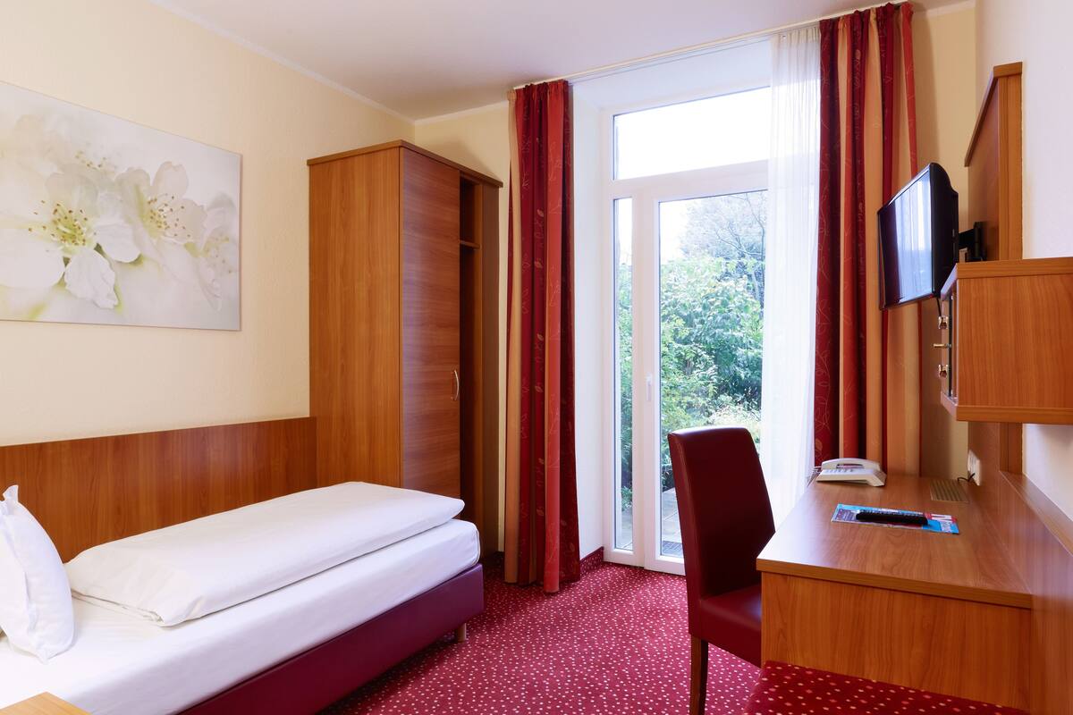 Quarto de hotel com cama, escrivaninha, televisão e janela com cortinas.