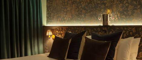 Quarto de hotel com cama, cortinas escuras, parede com papel de parede floral, abajur e minibar.