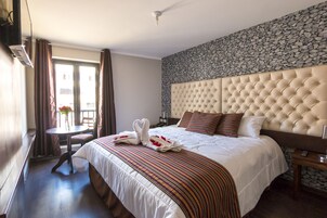 Quarto de hotel com uma cama grande, parede de pedra, uma mesinha e varanda com cortinas.