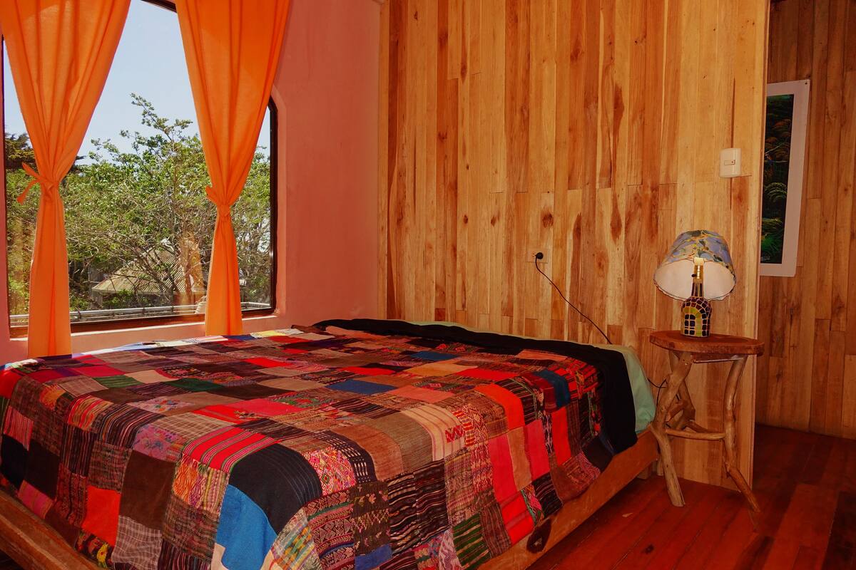 Quarto com parede de madeira, cama grande com colcha colorida, cortinas laranja e uma pequena mesa com luminária.