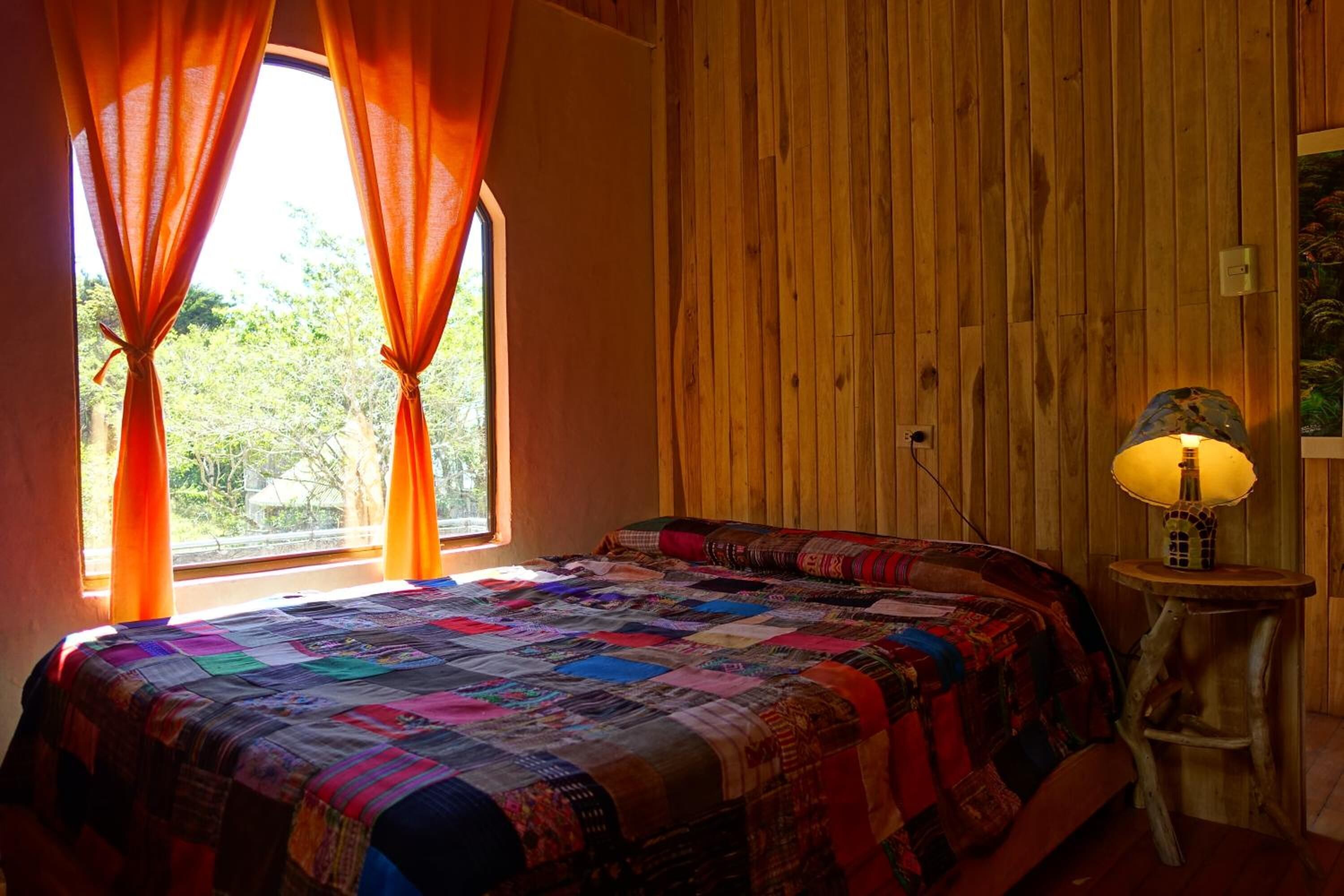 Quarto com parede de madeira, uma janela grande com cortinas laranja, cama com colcha colorida e abajur sobre um criado-mudo de madeira.