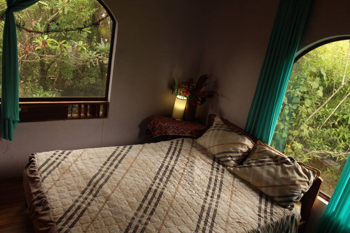 Um quarto com uma janela grande, uma mesa de cabeceira com abajur e uma cama com colcha acolchoada.