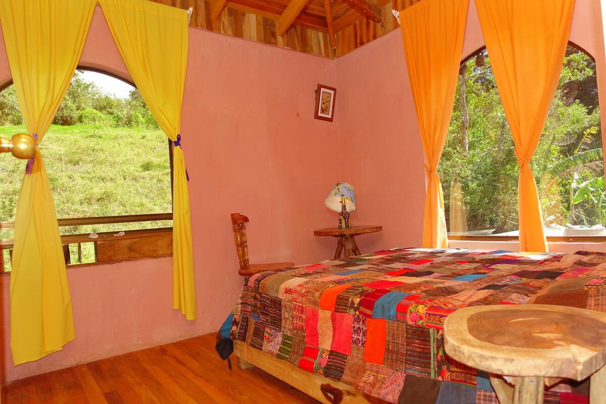 Quarto com cama de madeira, uma mesinha e cortinas.