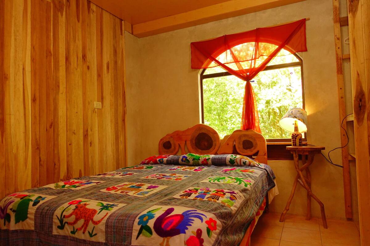 Quarto de madeira com cama, janela com cortina vermelha e cabeceira de madeira.