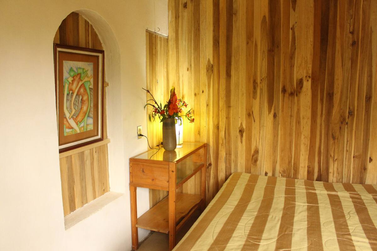 Quarto com paredes de madeira, cama com roupa de cama listrada, mesa de cabeceira de madeira com um vaso de flores e um quadro na parede.