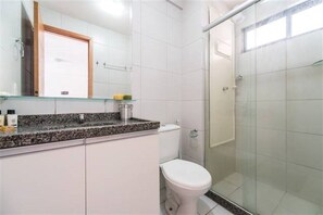 Casa de banho com cabine de duche em vidro, sanita branca e bancada de granito com espelho em cima.