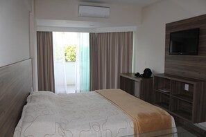 Quarto de hotel com uma cama, televisão fixada na parede, escrivaninha com cadeira e porta de vidro deslizante para uma varanda.
