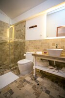 Um banheiro com chuveiro em cabine de vidro, vaso sanitário branco, pia com espelho acima e piso de azulejos.