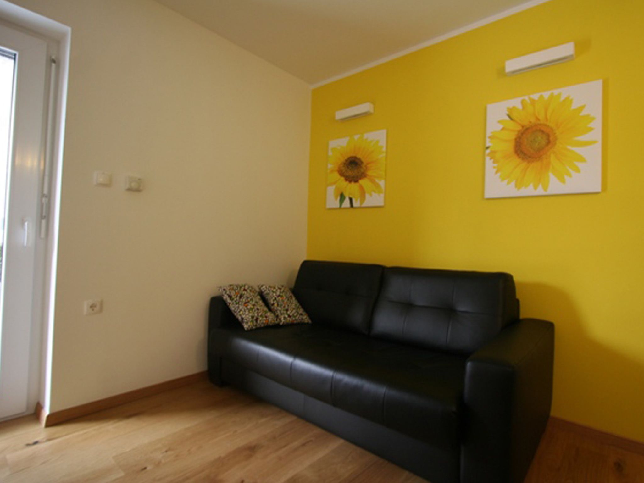 Quarto com sofá de couro preto, piso de madeira e duas pinturas de girassóis na parede amarela.