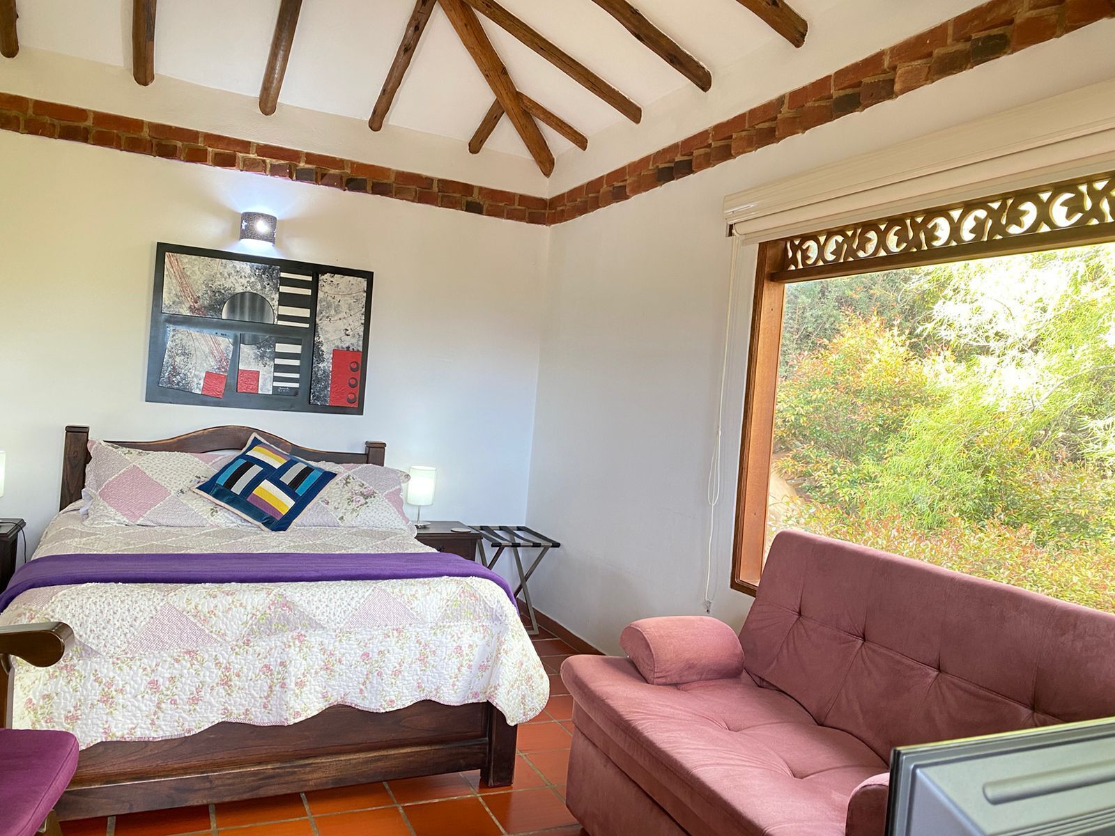 Quarto com cama, uma cadeira rosa, janela com vista para vegetação e um elemento decorativo na parede.
