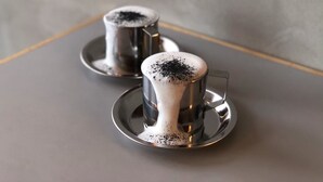 Duas chávenas de metal com pires, cada uma com uma bebida espumosa polvilhada com um pó escuro, provavelmente café ou cacau, sobre uma superfície reflectora.