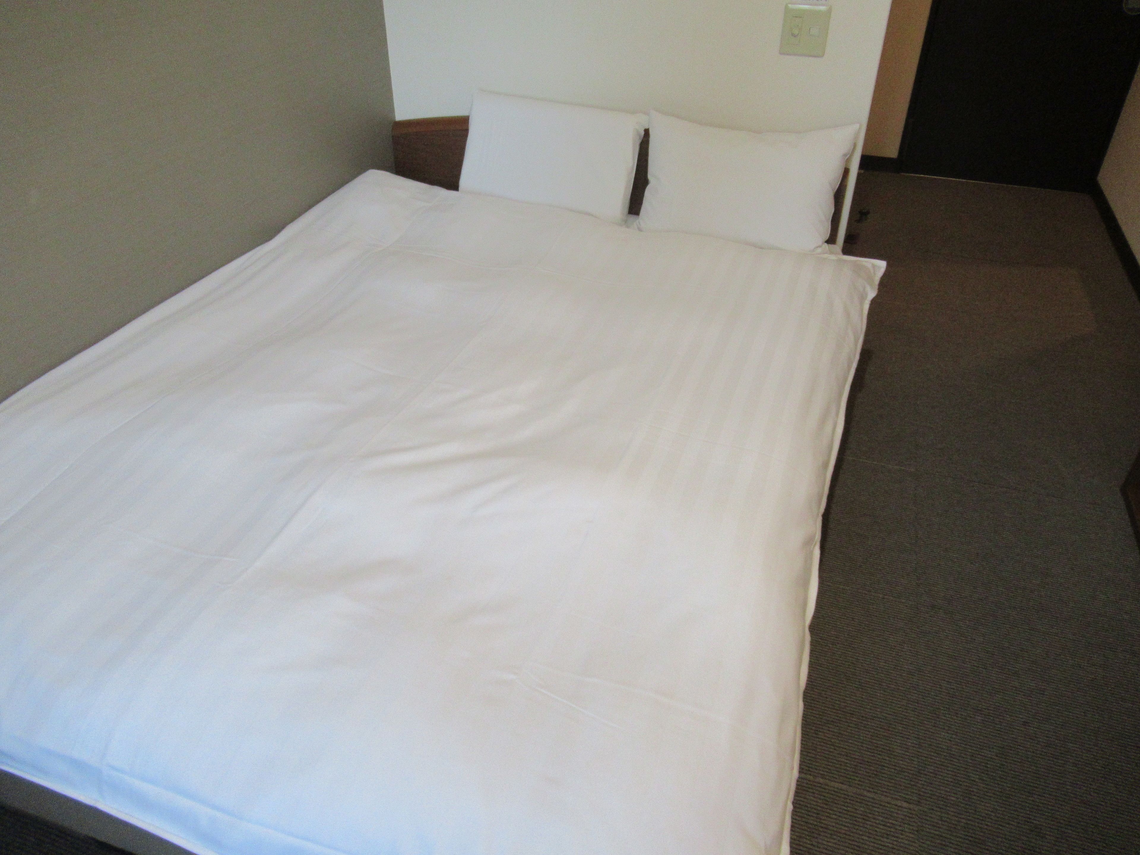 Uma cama de solteiro com roupa de cama branca num quarto com chão coberto de carpete e uma parede lisa.