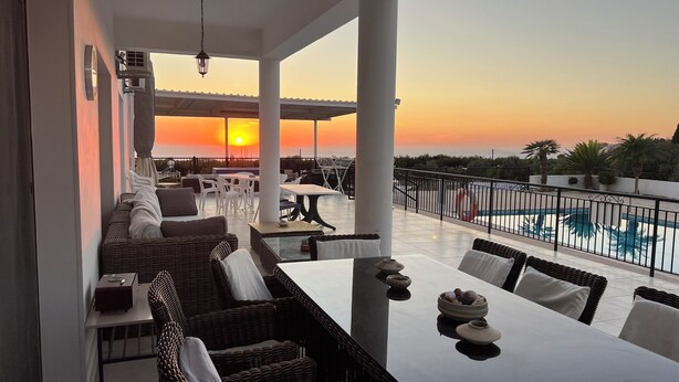 Terraço com área de refeições, mobiliário de vime e vista para o pôr do sol sobre o mar.